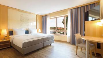 Quarto de hotel com uma cama grande, escrivaninha e uma cadeira. Há uma janela com cortinas e vista para edifícios.