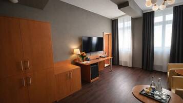 Quarto de hotel com um guarda-roupa de madeira grande, uma televisão de tela plana, uma escrivaninha com luminária, uma pequena mesa com bandeja e uma janela ampla com cortinas.