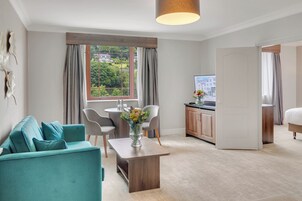 Quarto de hotel moderno com um sofá azul-esverdeado, mesa de centro de madeira, área de jantar com cadeiras, cozinha e uma janela grande com cortinas.