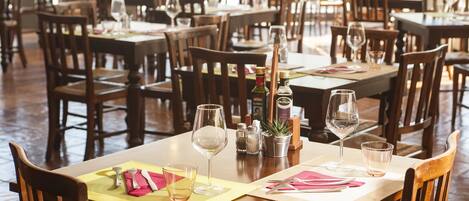 Restaurante com mesas e cadeiras de madeira, preparado com copos de vinho, garrafas e acompanhamentos.