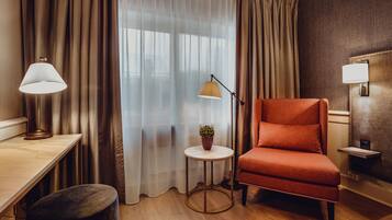 Quarto de hotel com piso de madeira, uma mesa redonda, uma poltrona vermelha, uma pequena mesinha de cabeceira com abajur e um vaso de planta.