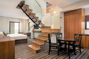 Quarto de hotel moderno com escada de madeira, área de refeições com mesa redonda e cadeiras, cama com cabeceira e um guarda-roupa grande de madeira.