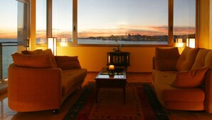Sala de estar com dois sofás, uma mesa de centro e vista para o horizonte urbano ao pôr do sol.