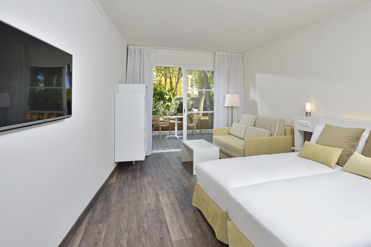 Quarto de hotel com cama, sofá, mesinha, televisão e porta de correr de vidro que dá para uma área externa.