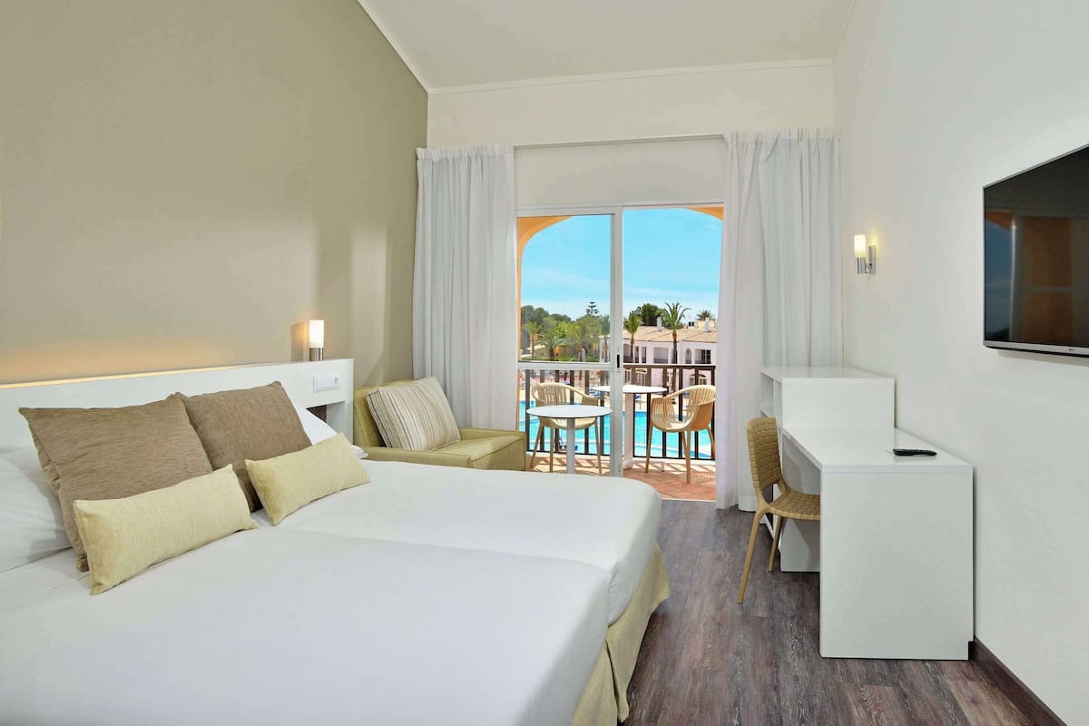 Quarto de hotel com uma cama grande, escrivaninha, televisão e varanda com piscina.