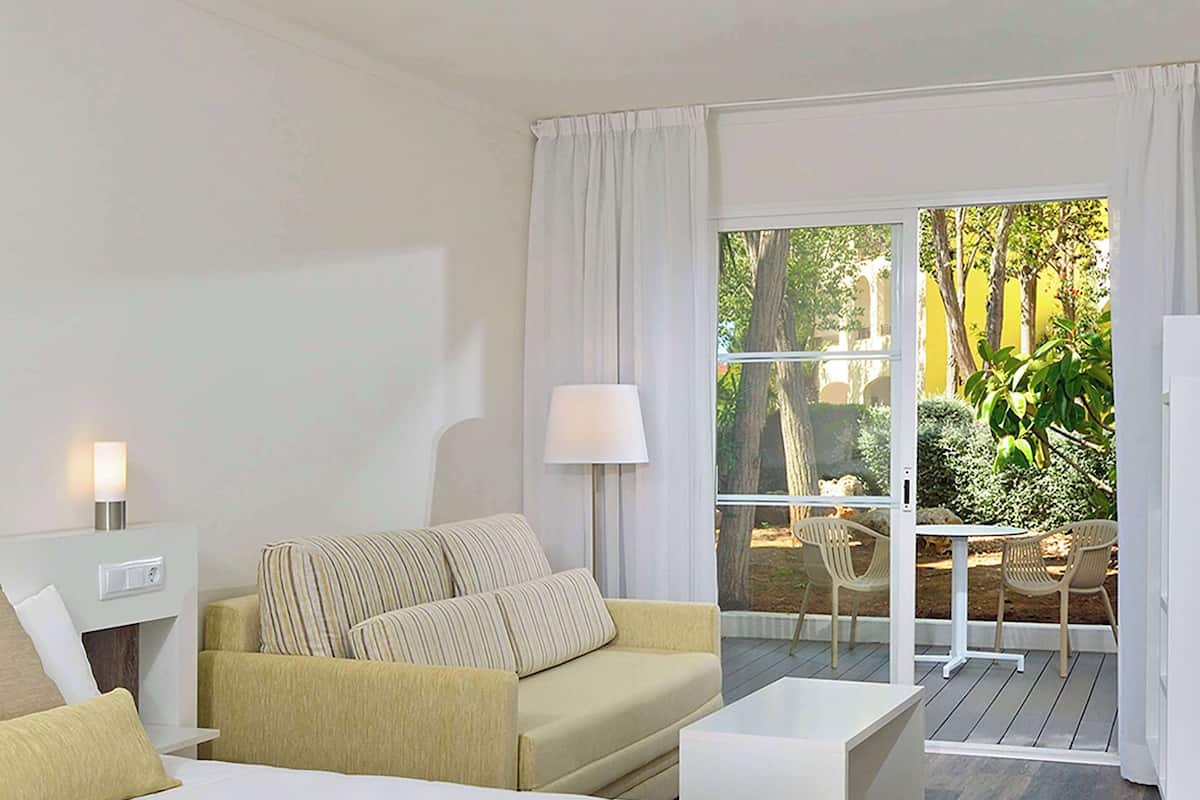 Quarto de hotel com uma cama grande, um sofá, uma pequena mesa e vista para a área externa por uma porta de vidro.