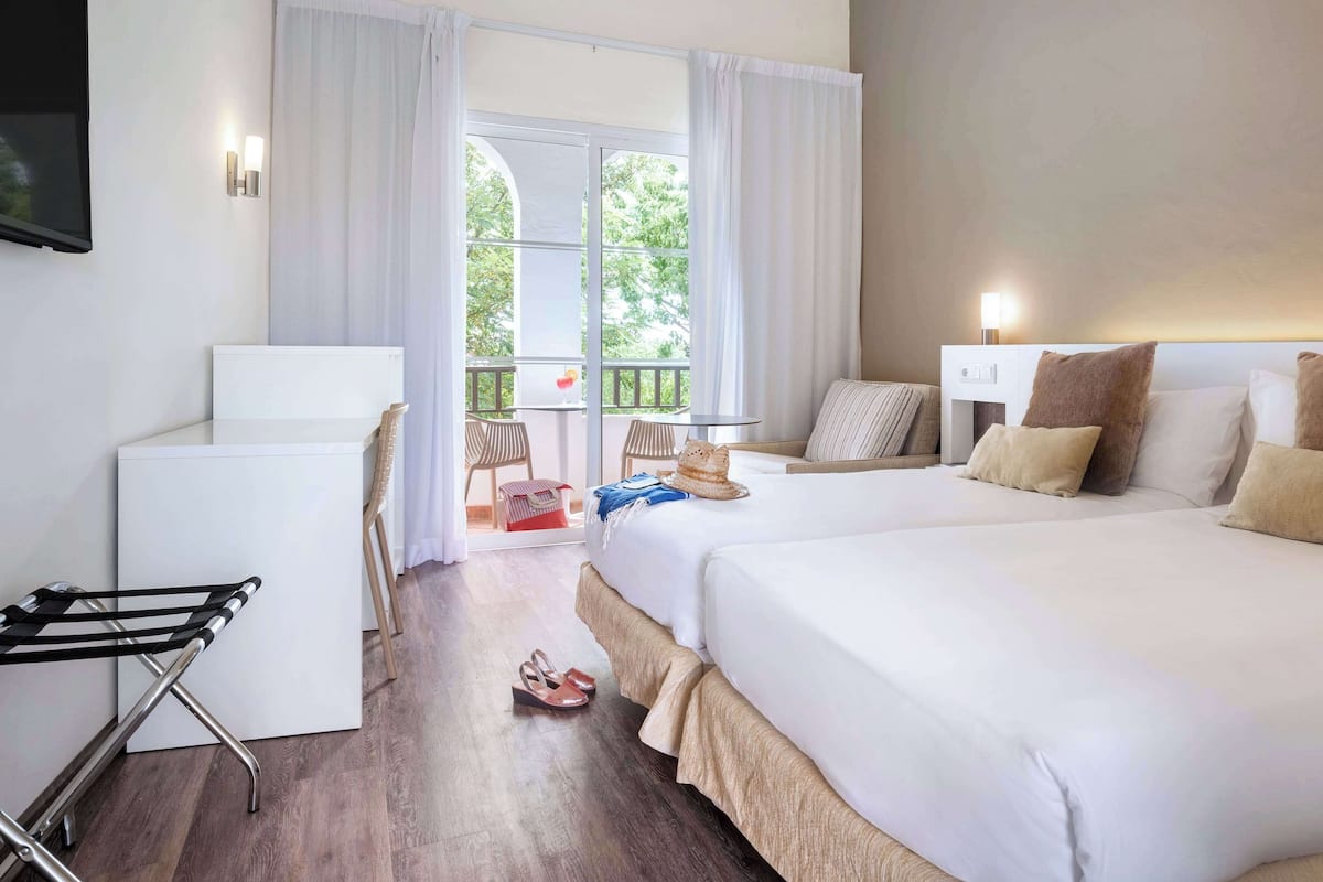 Quarto de hotel com duas camas, uma escrivaninha, uma cadeira e varanda com vista para o verde.