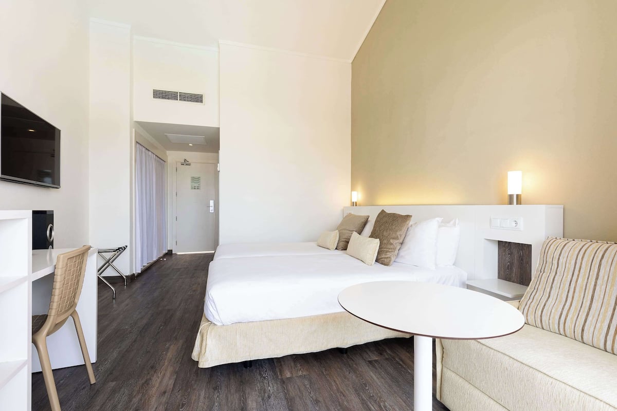 Quarto de hotel moderno com uma cama grande, um sofá pequeno, uma mesa redonda e uma TV fixada na parede.
