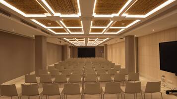Uma sala de conferências com fileiras de cadeiras, uma tela grande e um teto com iluminação geométrica.
