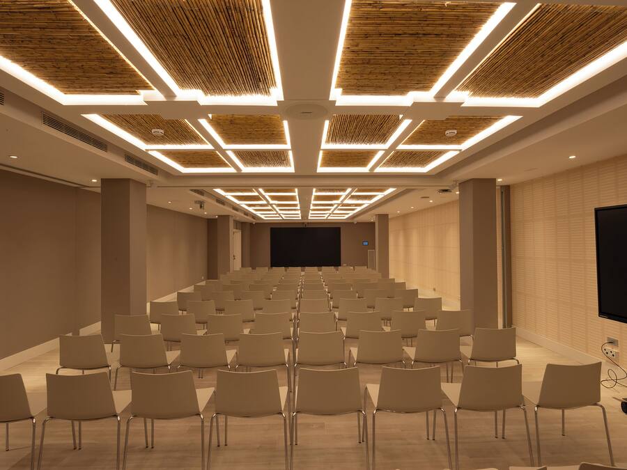 Uma sala de conferências com fileiras de cadeiras, uma tela grande e um teto com iluminação geométrica.