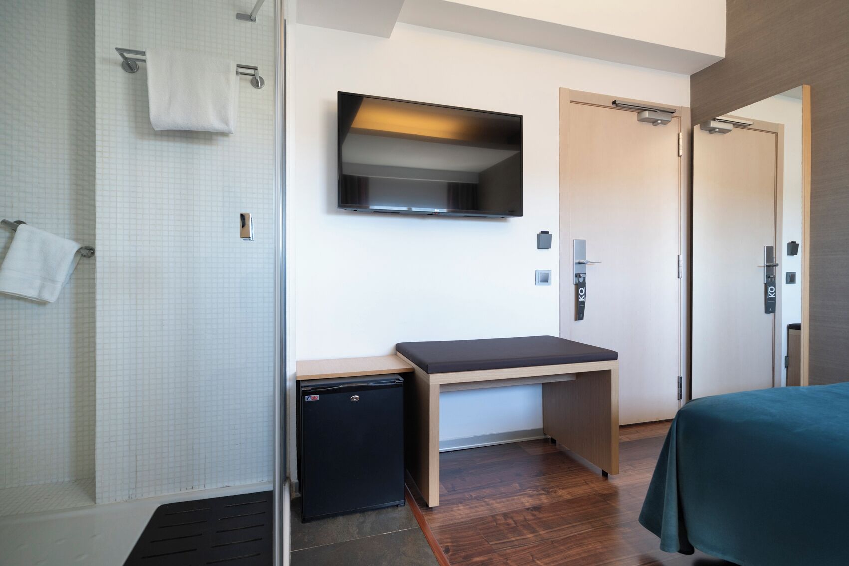 Quarto de hotel compacto com uma pequena mesa, televisão montada acima, mini-geladeira e chuveiro.