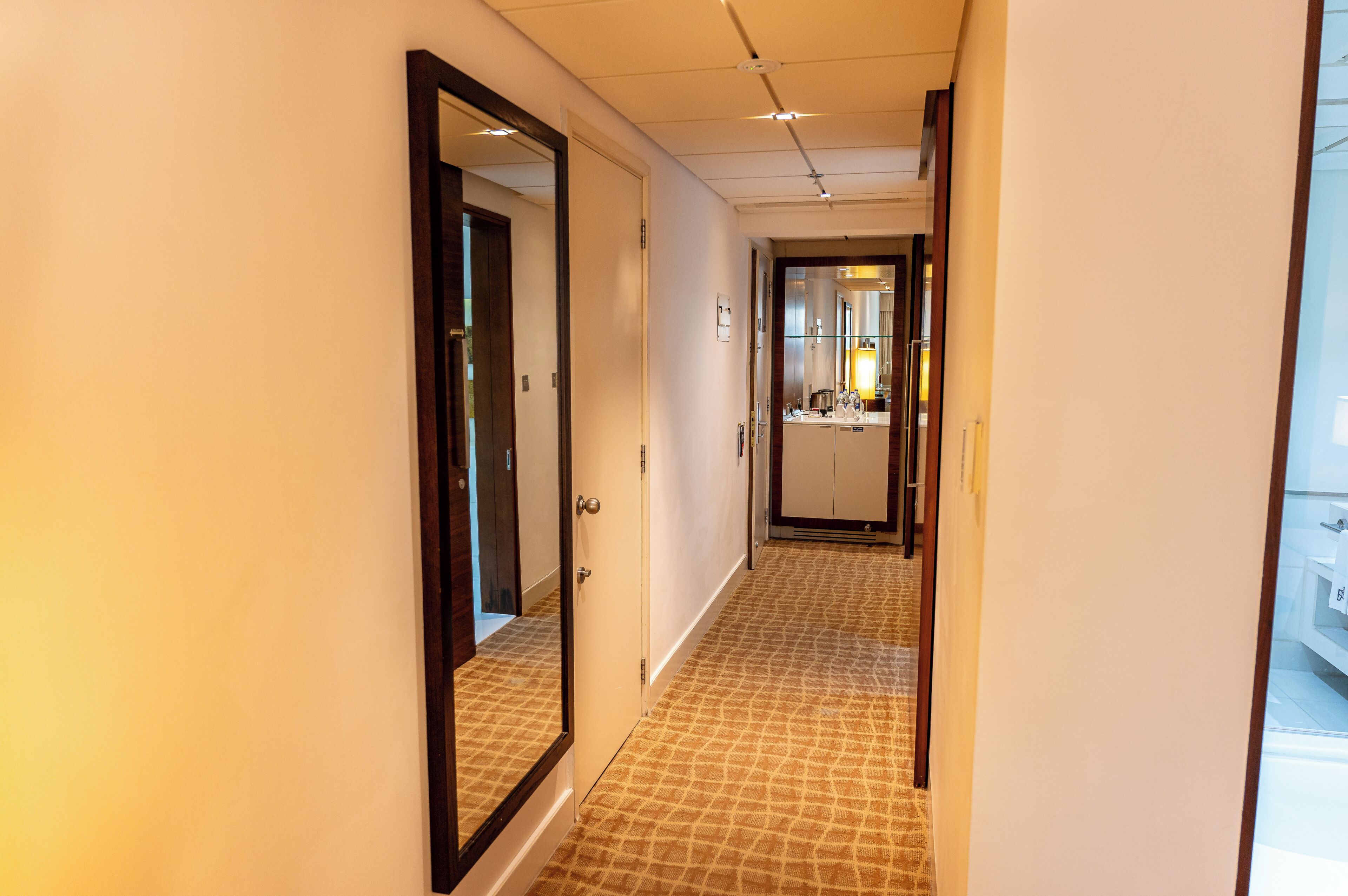 Corredor de hotel com piso estampado, um espelho à esquerda e vista para um quarto com pia e espelho.