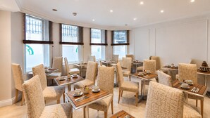 Uma sala de jantar de hotel com várias mesas e cadeiras, janelas amplas com persianas e um interior moderno e impecável.