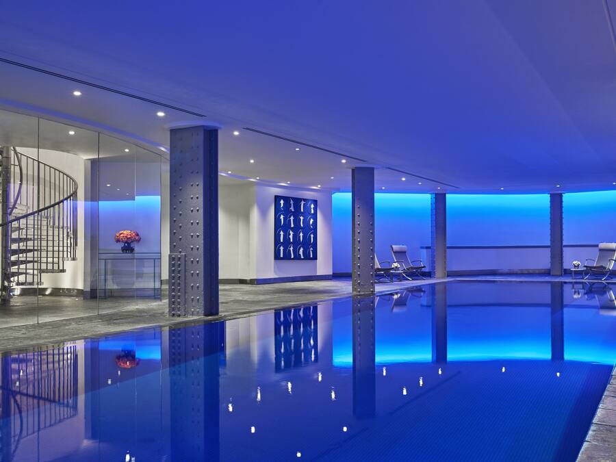 Uma piscina interior moderna com iluminação azul translúcida, uma escadaria em espiral e espreguiçadeiras.