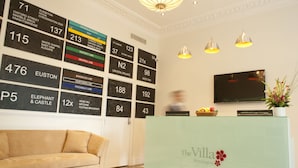 Uma área de recepção de hotel com piso xadrez, um sofá, um balcão de recepção com a placa 'The Villa Kensington' e uma parede com placas de quarto numeradas.