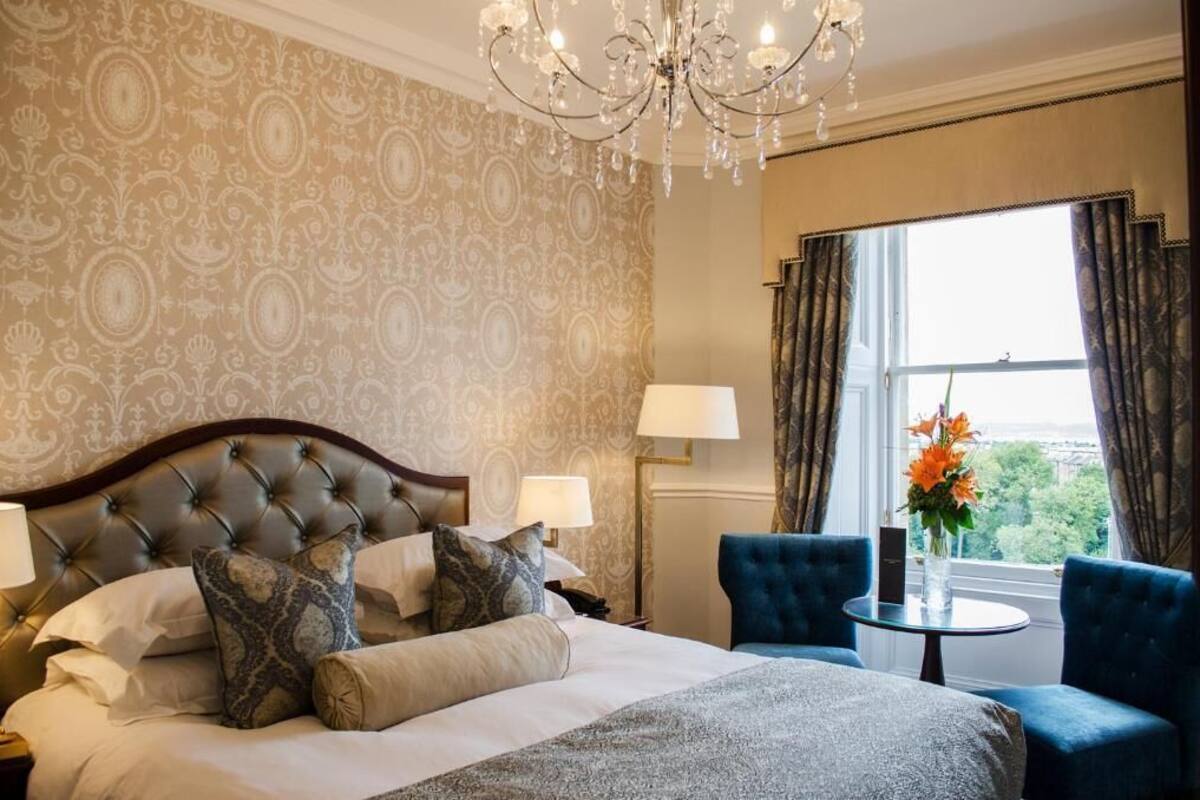 Quarto de hotel com cabeceira estofada, cama com almofadas estampadas, mesa redonda com um vaso de flores e vista para o exterior através de uma janela com cortinas.
