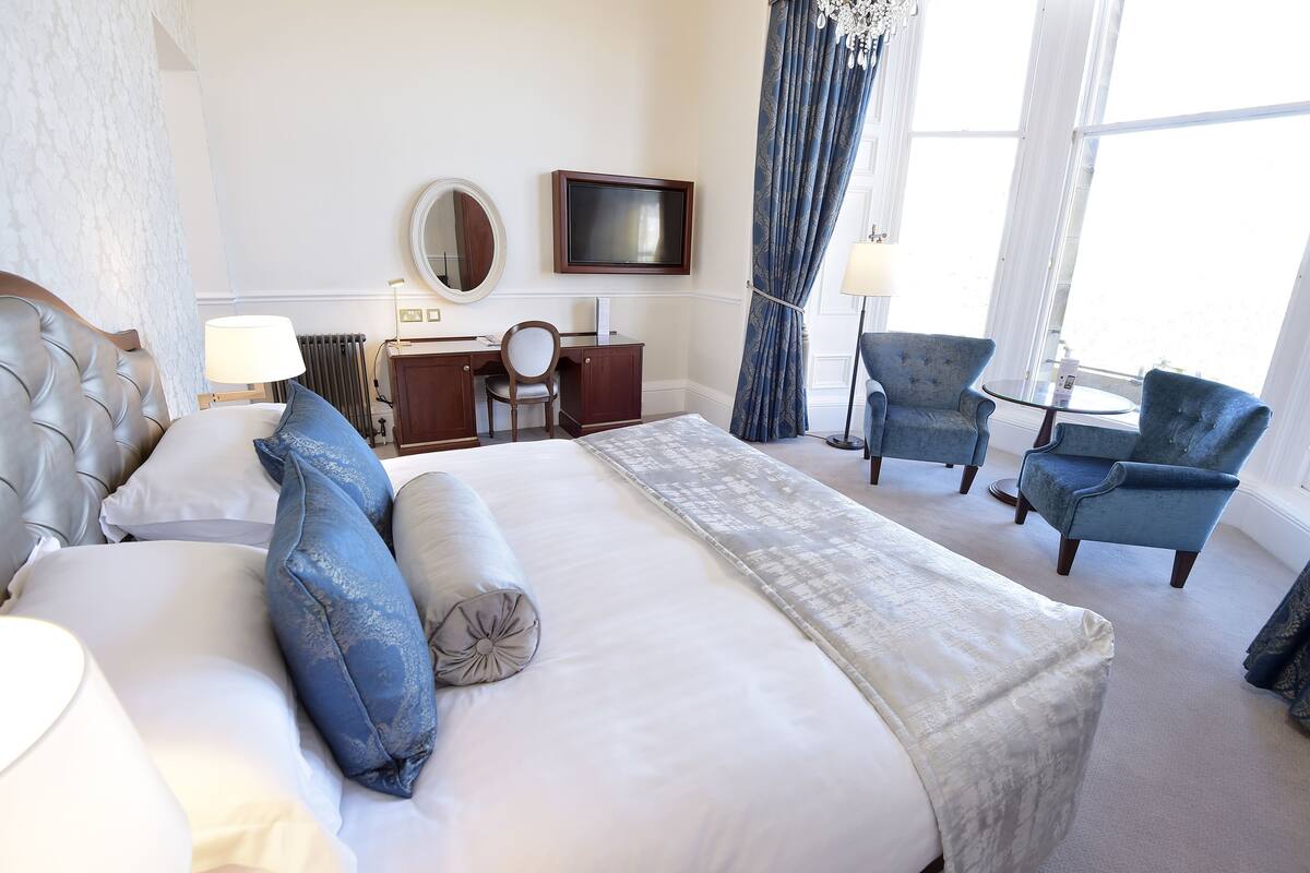 Quarto de hotel com uma cama grande, uma escrivaninha com cadeira, televisão e um espaço de estar com cadeiras.