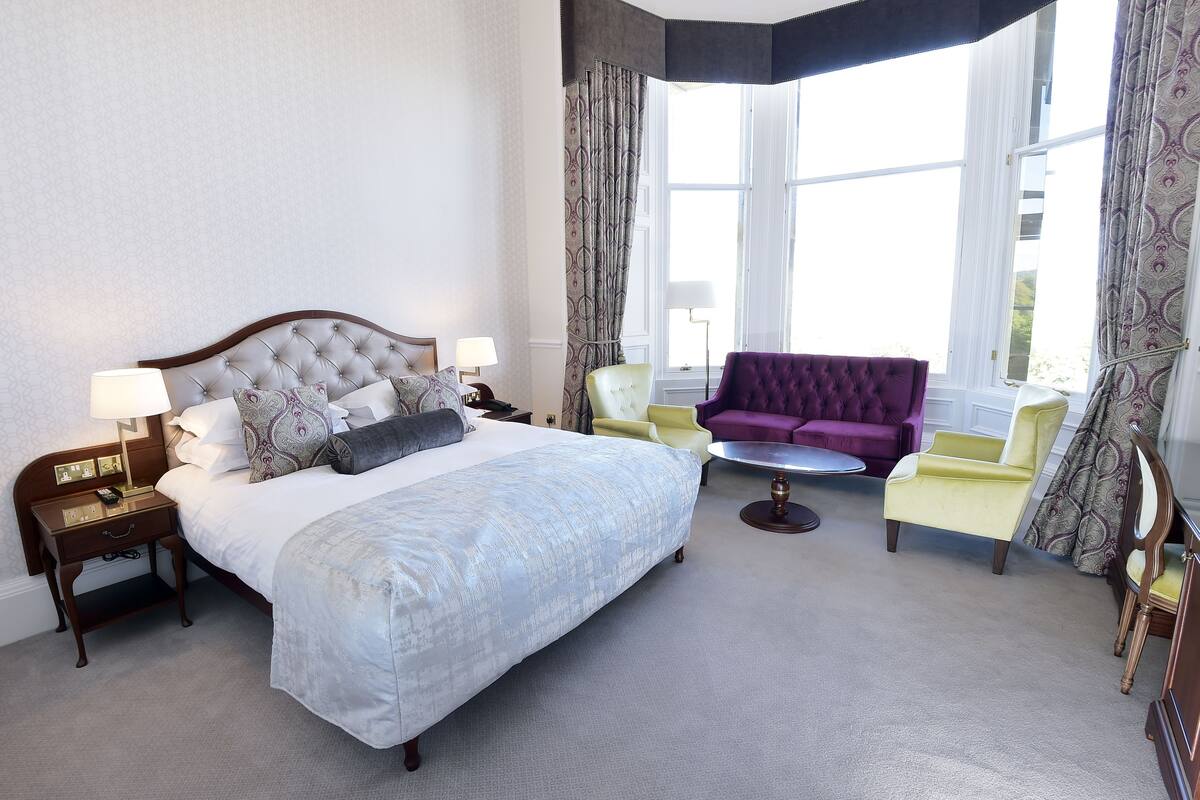 Quarto de hotel com uma cama grande, uma mesa de cabeceira, uma mesinha de apoio, uma poltrona roxa e uma poltrona amarela, além de uma pequena mesa redonda.