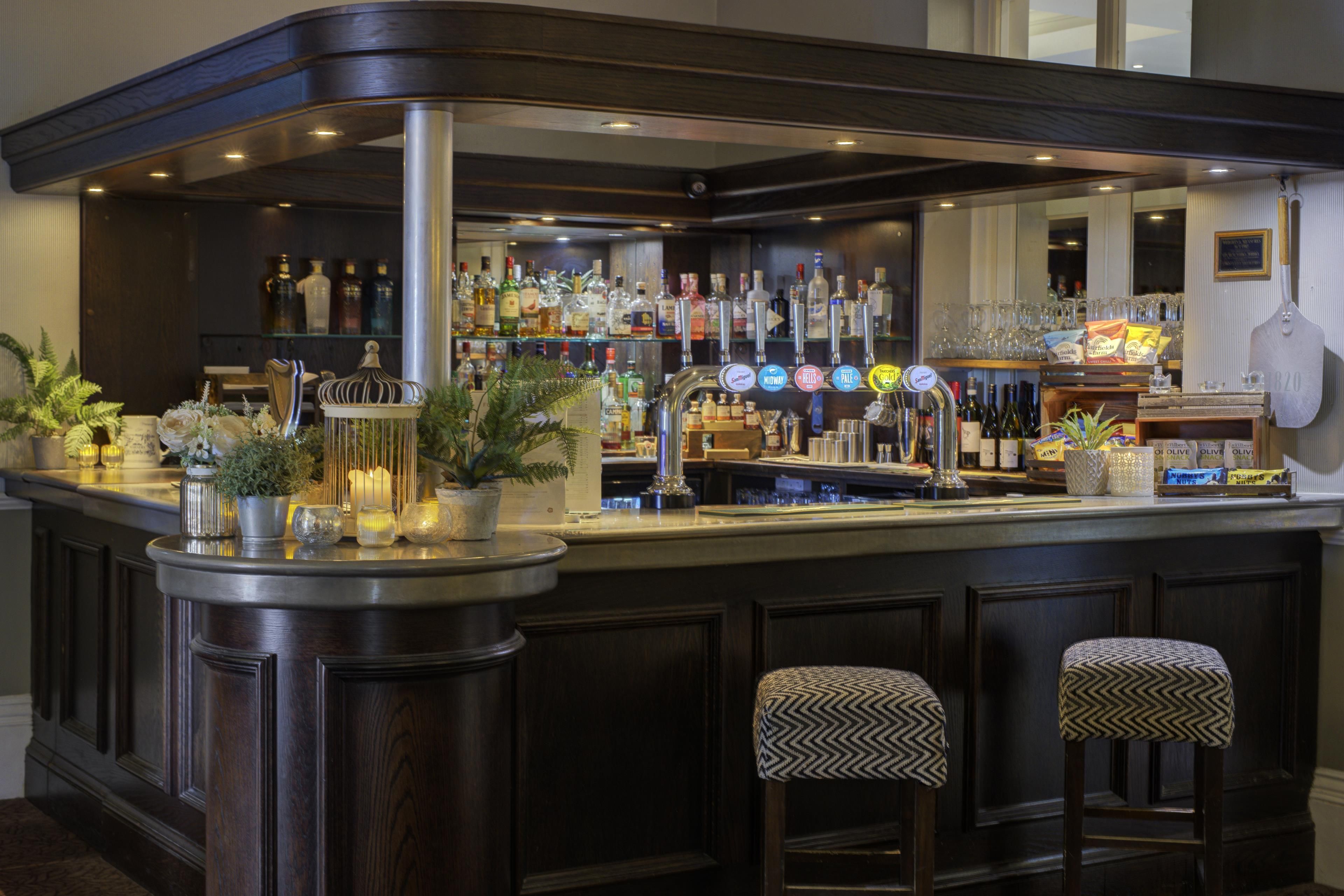 Uma área de bar com diversas garrafas de bebidas, vários tipos de copos e um grande cuspidor.
