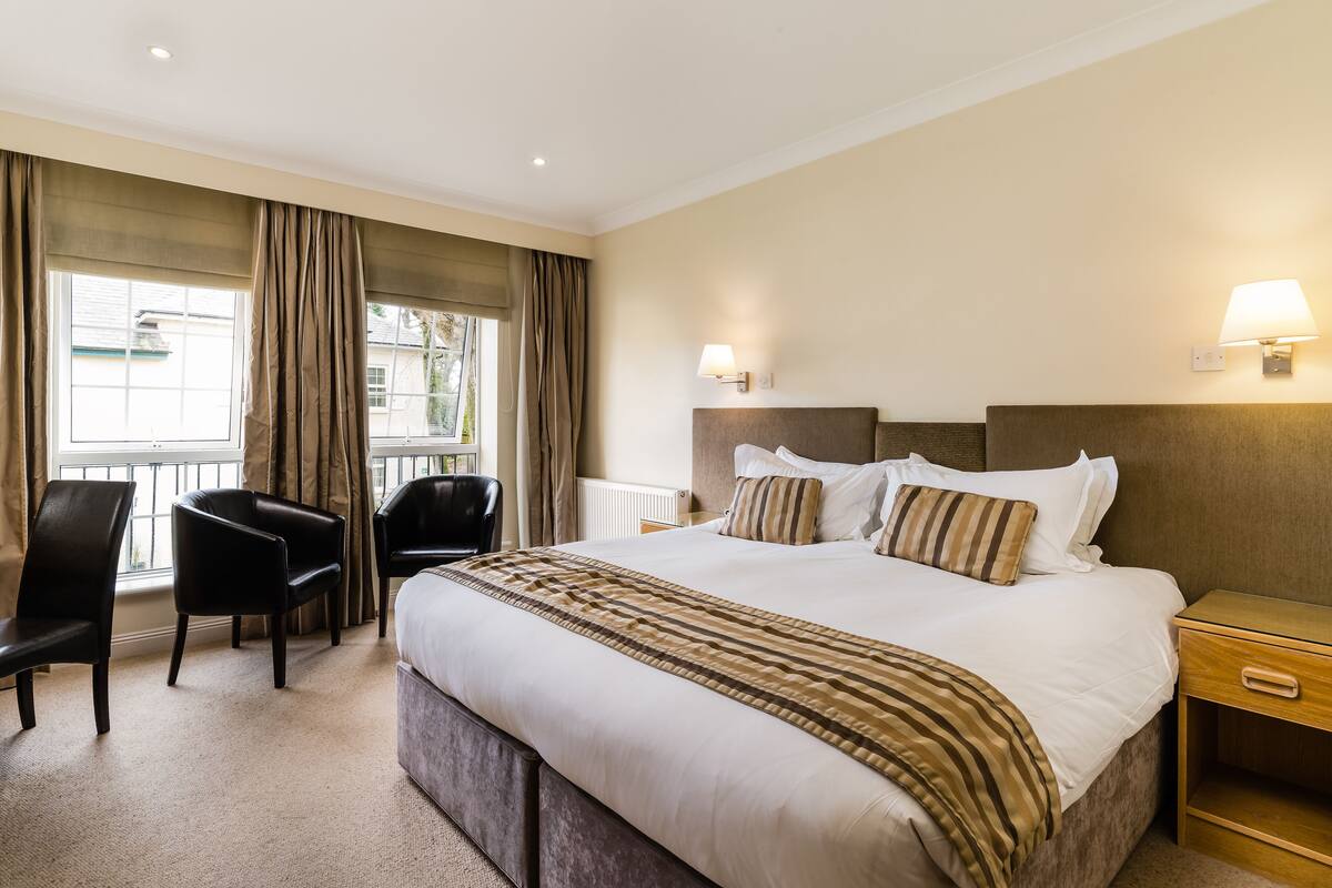 Quarto de hotel com uma cama grande, duas mesas de cabeceira, uma cadeira e vista para o exterior.