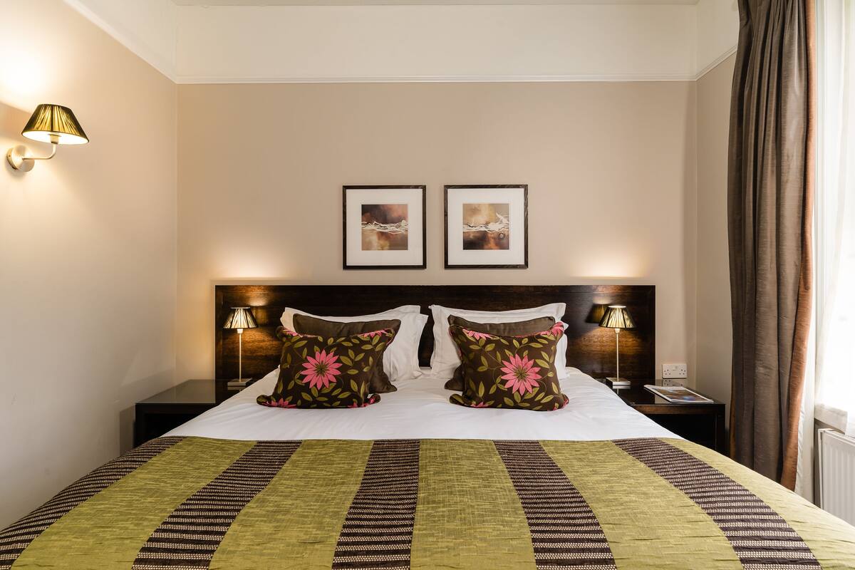 Uma cama bem arrumada com um cobre-leito estampado, duas luminárias fixadas na parede e duas molduras com quadros.