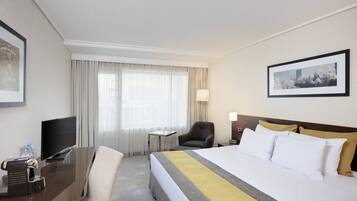 Quarto de hotel com cama, escrivaninha com televisão, cadeira, abajur e quadro pendurado na parede.