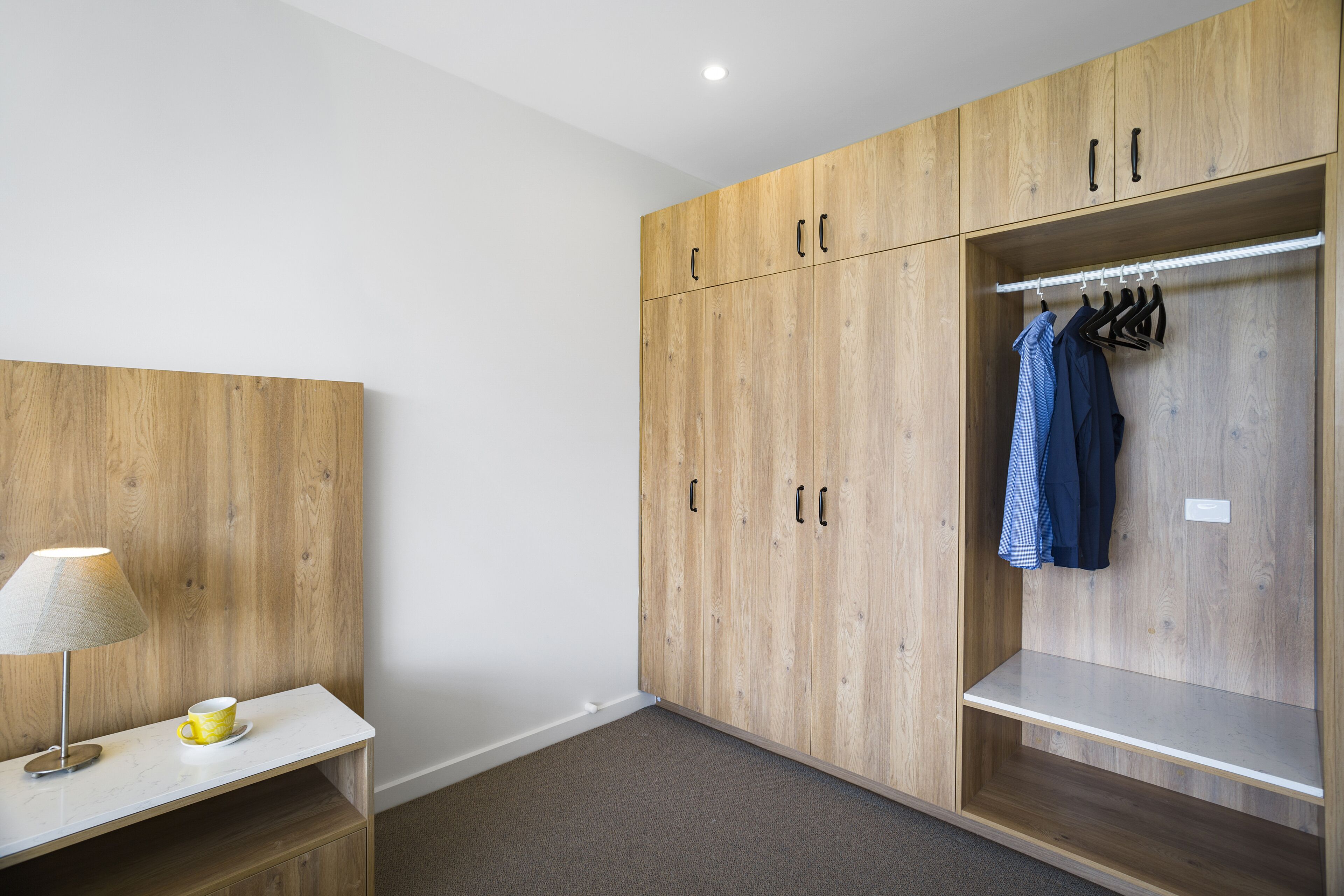 Quarto com guarda-roupa de madeira, uma mesa pequena com luminária e um cabideiro com roupas.