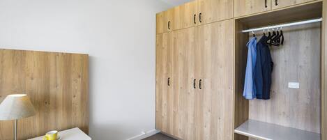 Quarto com guarda-roupa de madeira, uma mesa pequena com luminária e um cabideiro com roupas.