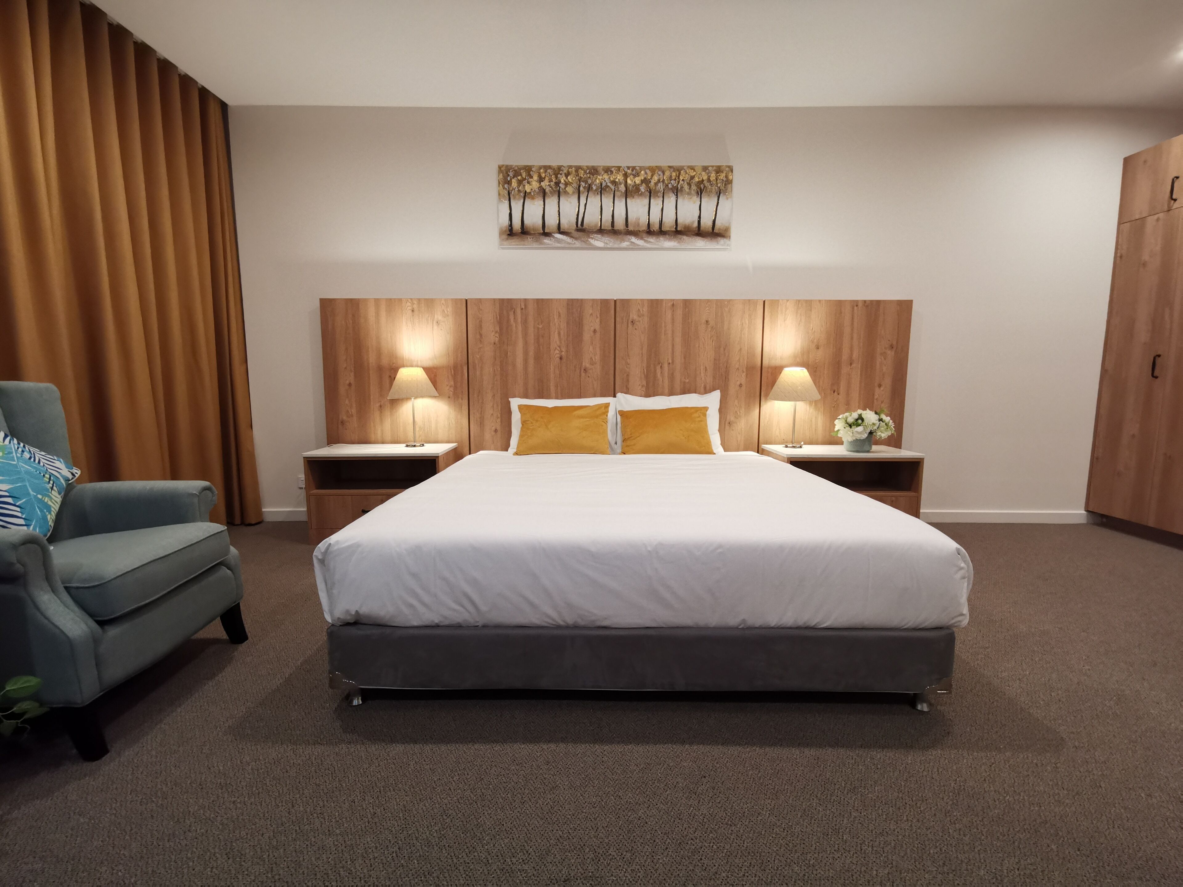Quarto de hotel moderno com cama grande, duas mesinhas de cabeceira com luminárias, cabeceira de madeira, poltrona azul e uma obra de arte na parede.