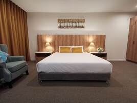 Quarto de hotel moderno com cama grande, duas mesinhas de cabeceira com luminárias, cabeceira de madeira, poltrona azul e uma obra de arte na parede.