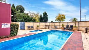 Piscina com placa de aviso, rodeada por um terraço de tijolos, área de estar ao ar livre e cerca de madeira.