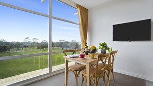 Uma área de refeições com mesa, cadeiras, uma televisão e vista para uma paisagem verdejante por amplas janelas.
