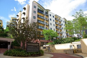 Um edifício residencial com vários andares, varandas, um aviso indicando comodidades como um restaurante no pátio e estacionamentos, e vegetação exuberante ao redor da propriedade.
