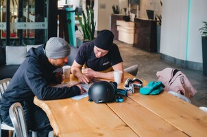 Duas pessoas em uma mesa de madeira, com um mapa, um capacete e um par de sapatos.
