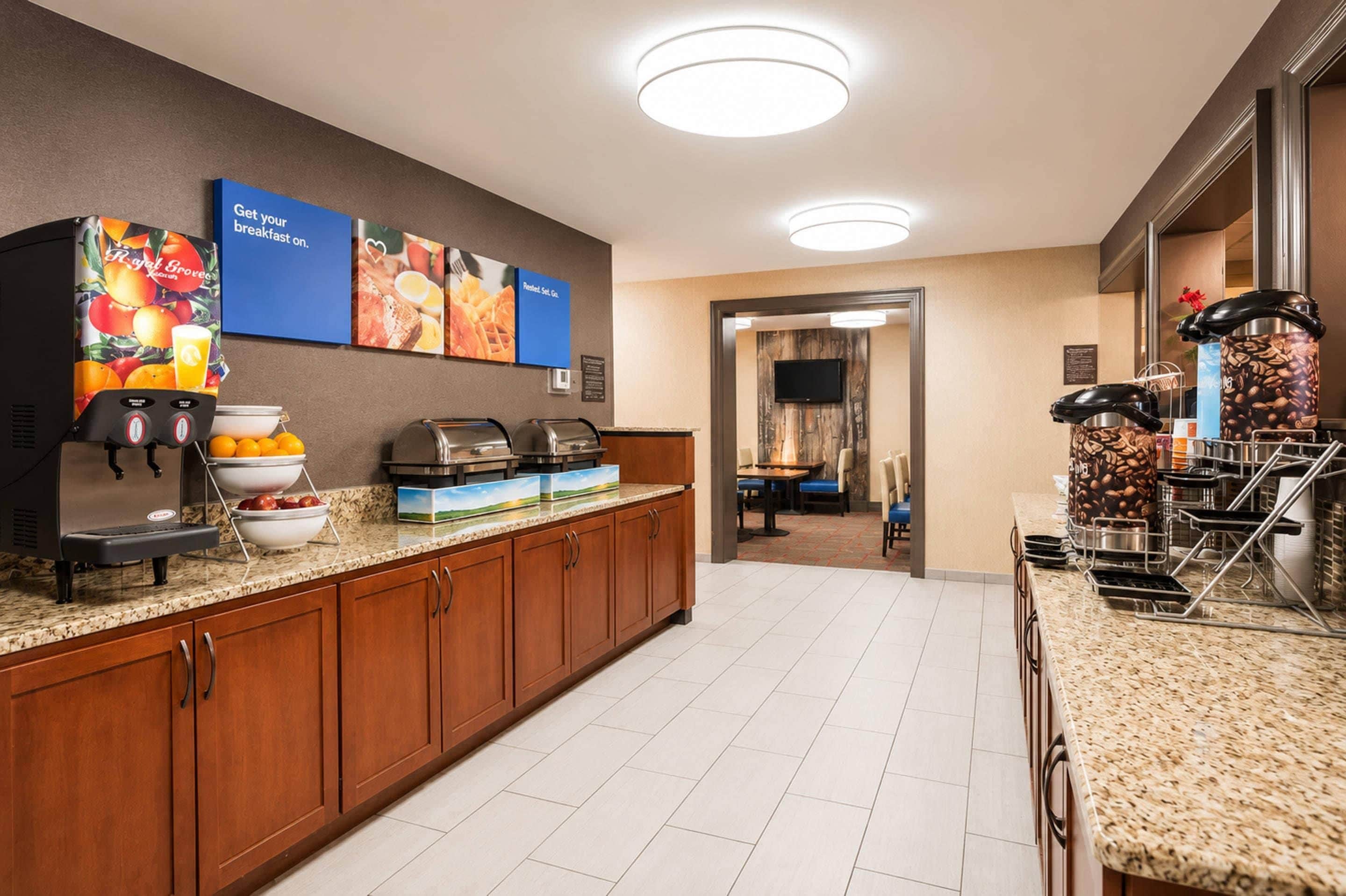 Espaço de café da manhã de hotel, com máquina de café, tigela de frutas e dispensador de café.