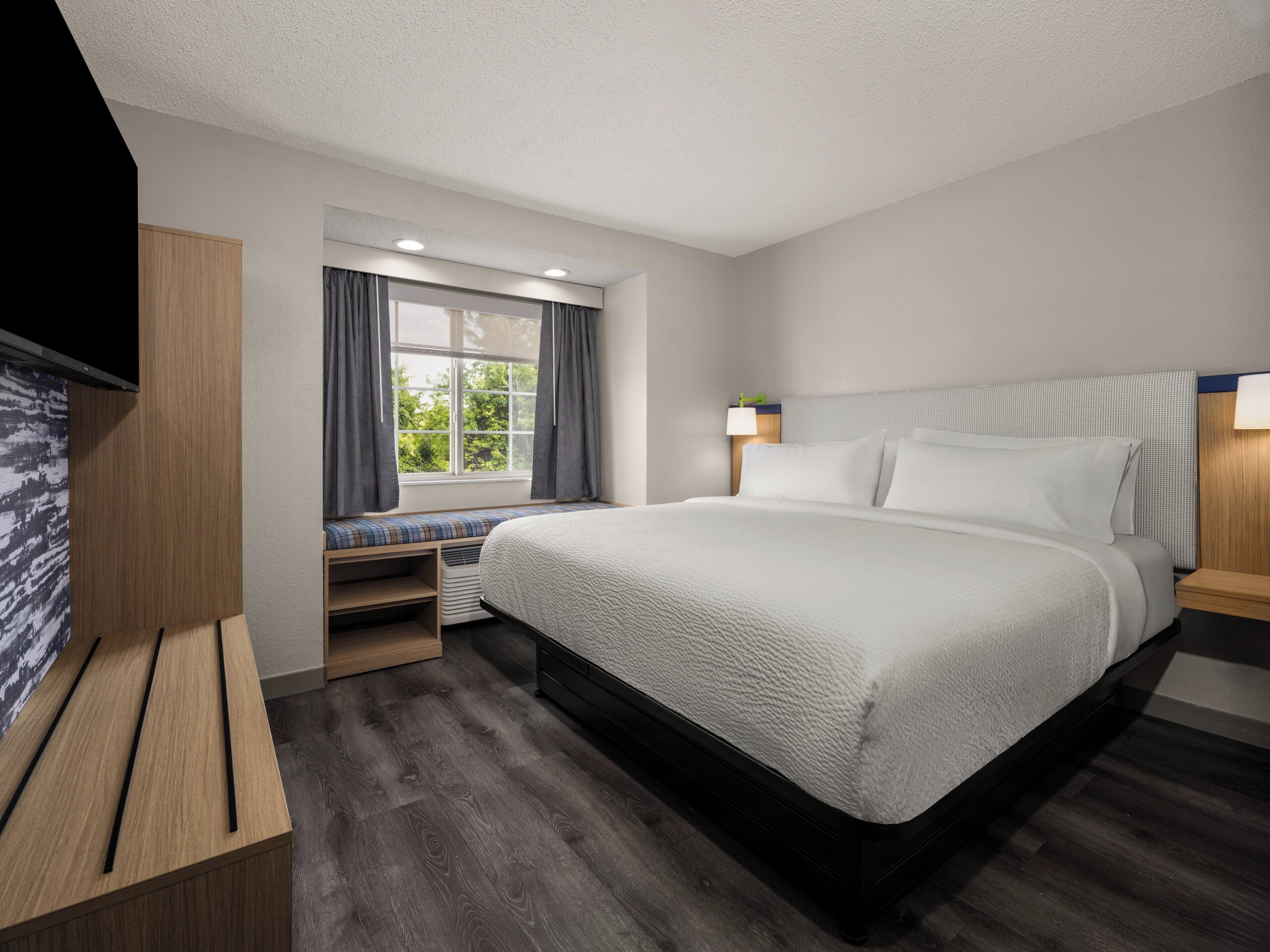 Quarto de hotel com uma cama grande, um banco com espaço de armazenamento, televisão e uma janela com cortinas.