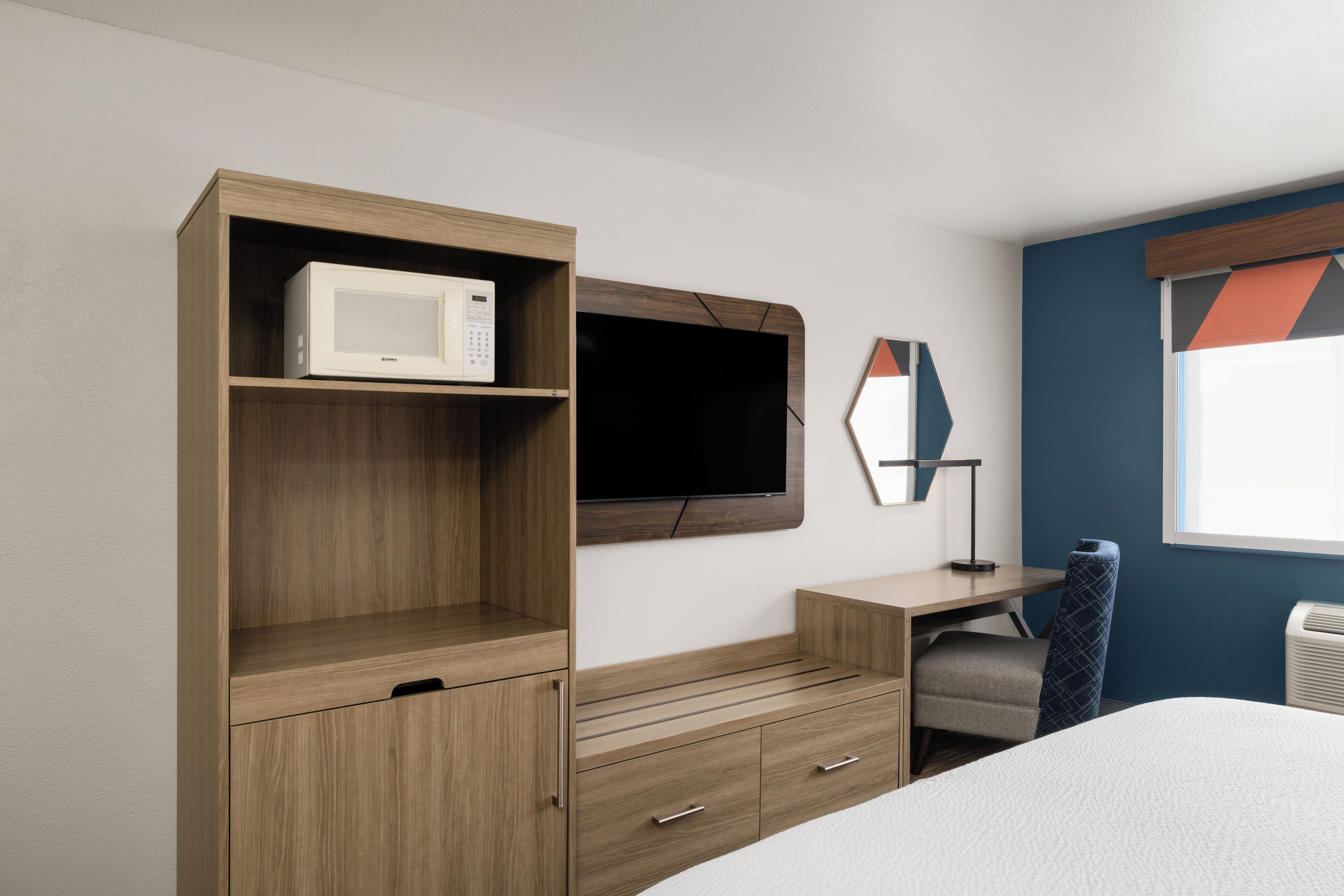 Um quarto com cama, escrivaninha com cadeira, guarda-roupa, televisão e janela com persianas.