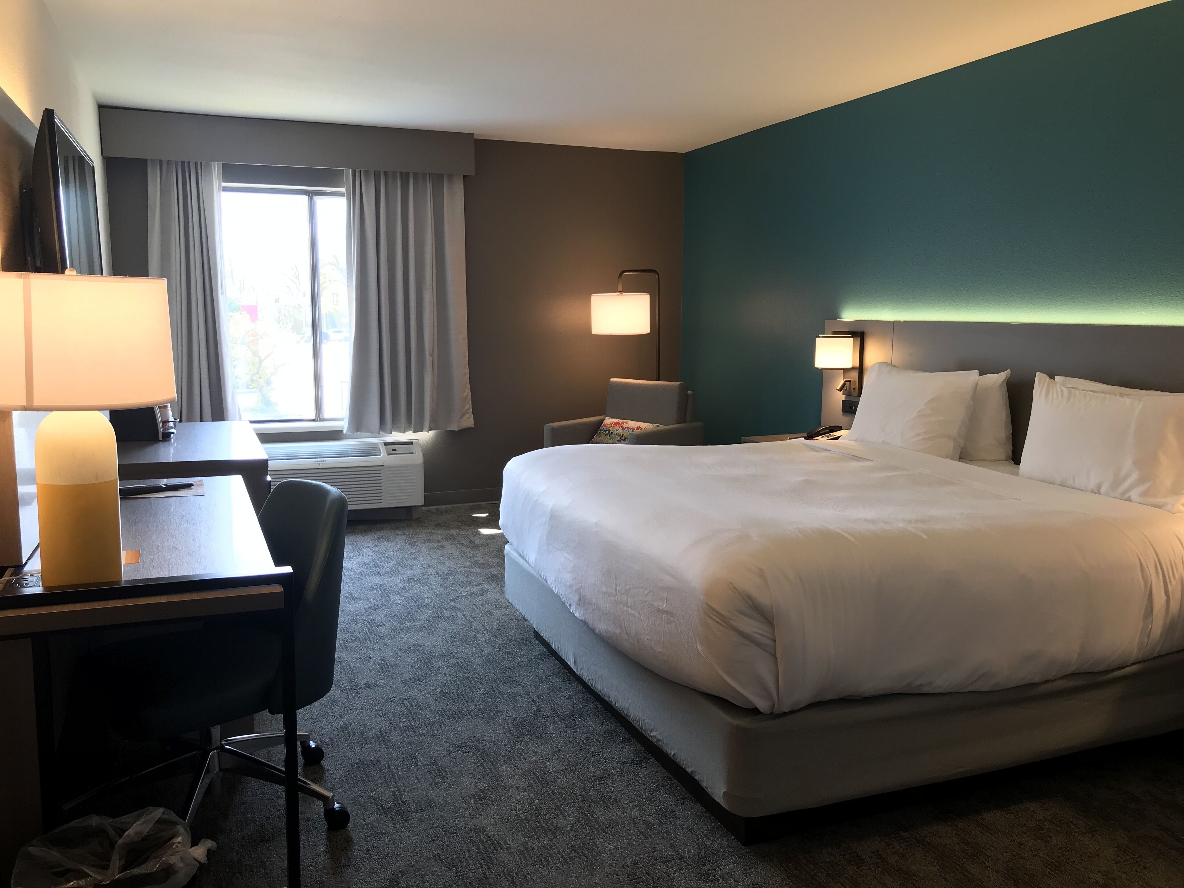 Quarto de hotel com cama, escrivaninha, cadeira e janela com cortinas.