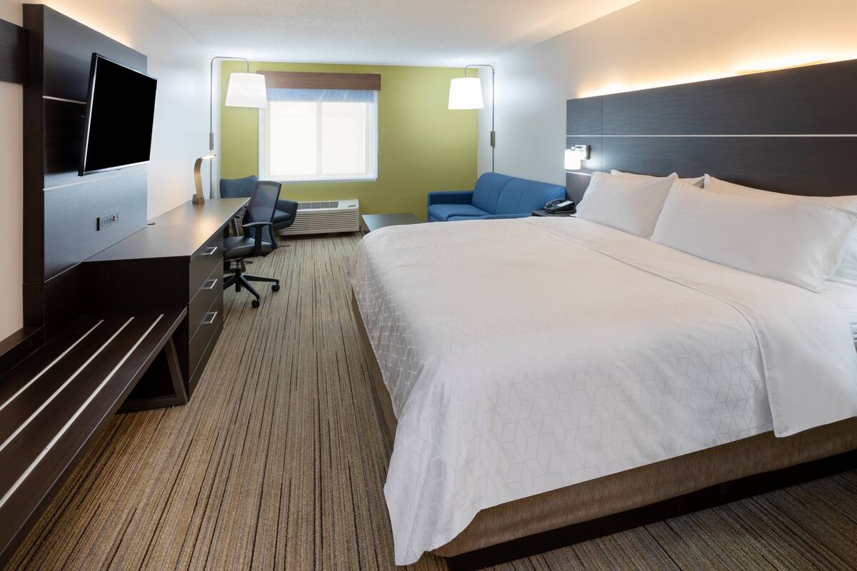 Quarto de hotel com uma cama grande, uma escrivaninha com computador, televisão e um espaço de estar com poltrona e luminária.