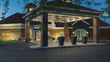 Entrada de hotel com galeria coberta, grandes vasos de plantas e uma placa bem visível.
