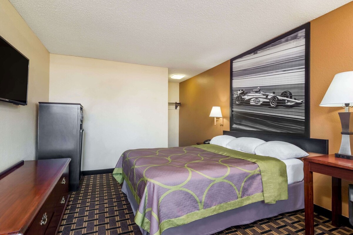 Quarto de hotel com cama, frigorífico, secretária, candeeiro e televisão montada na parede.