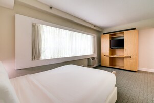 Quarto de hotel com uma cama, uma janela grande com cortinas, uma televisão sobre um suporte de madeira e piso coberto de carpete.