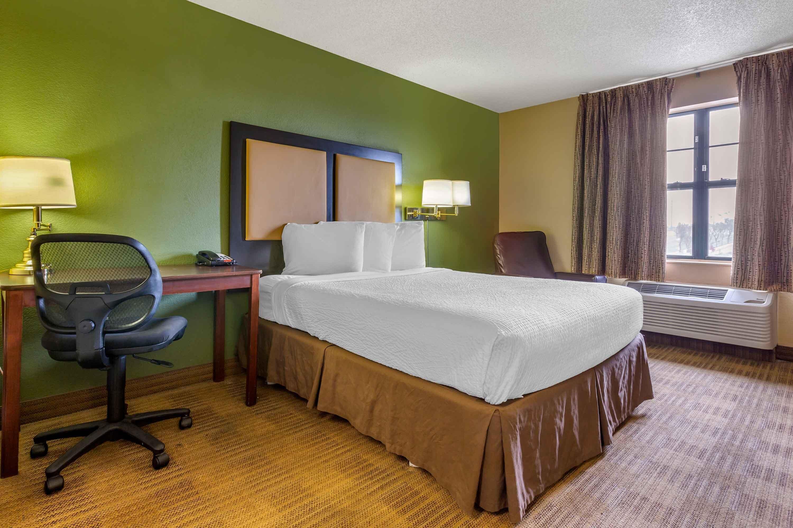 Quarto de hotel com cama, escrivaninha, cadeira de escritório, luminárias e ar condicionado.