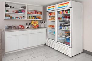 Uma cozinha com armários brancos, bancada com lanches e uma geladeira cheia de bebidas.