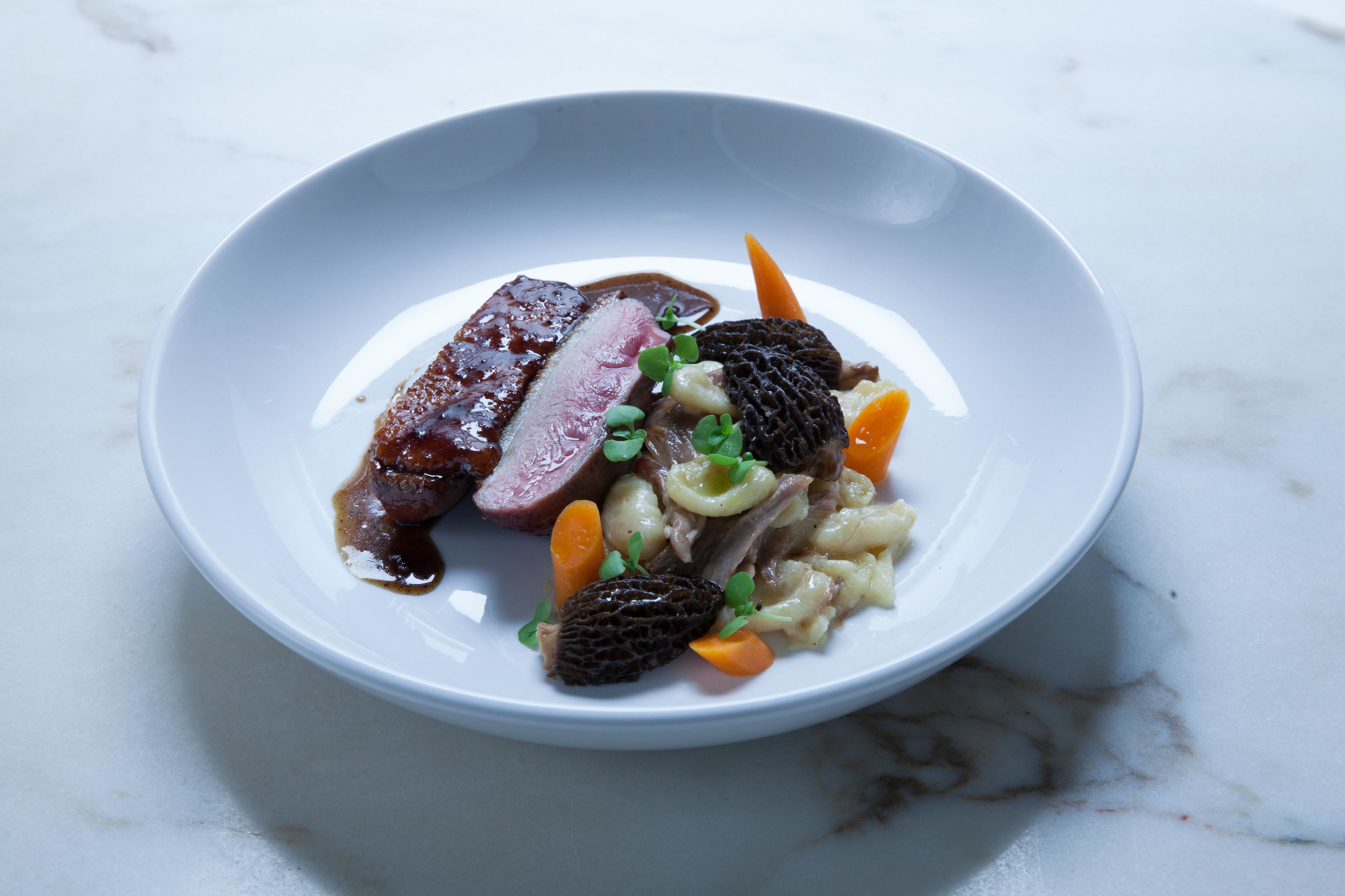 Um prato branco com um pedaço de carne, cenouras e um cogumelo como guarnição sobre uma superfície de mármore.