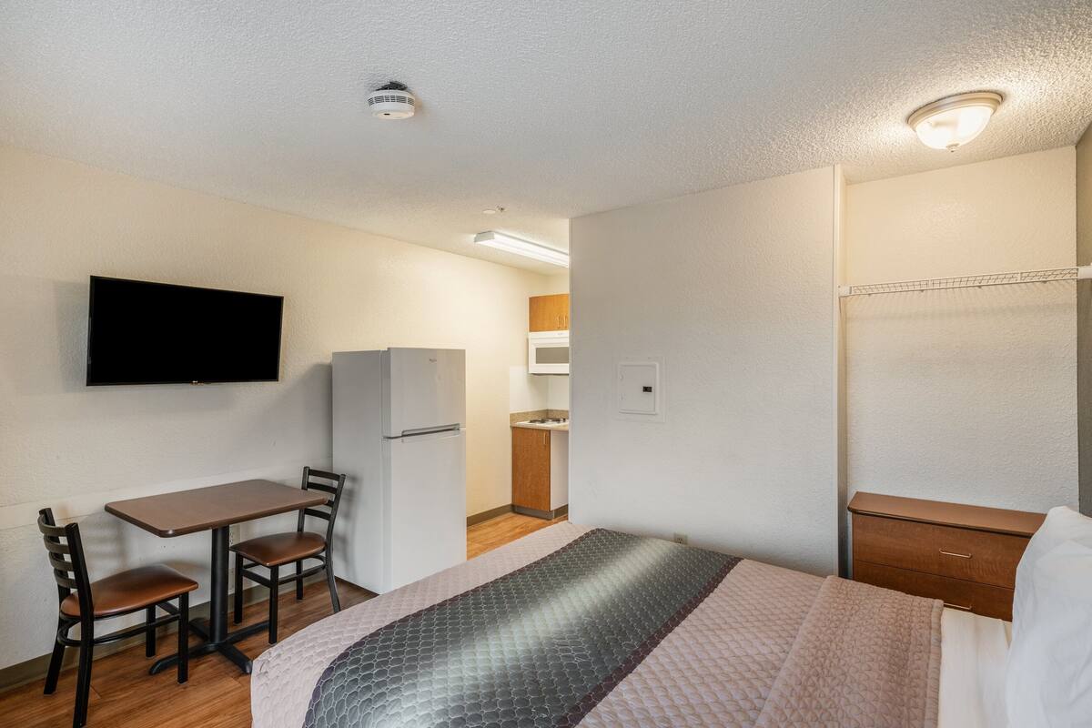 Quarto de hotel compacto com cama, escrivaninha com cadeiras, geladeira, televisão e pequena cozinha.