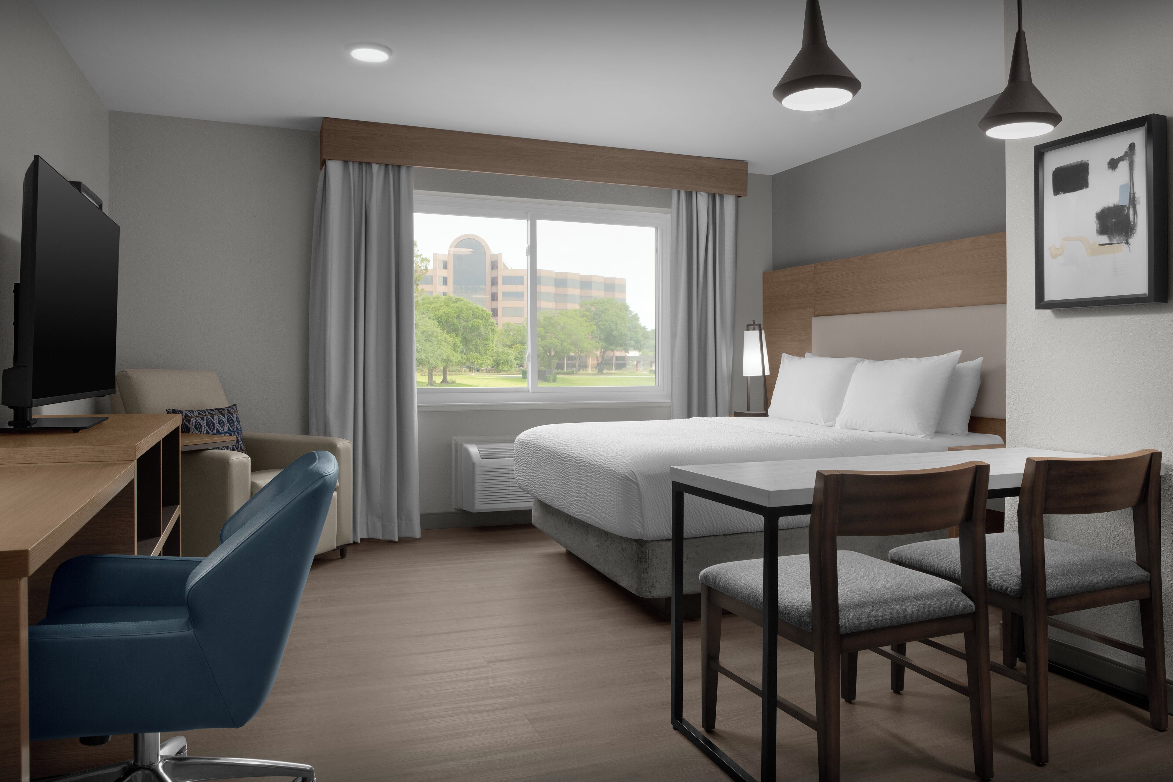 Quarto de hotel com cama, escrivaninha, cadeira e vista para um edifício pela janela.