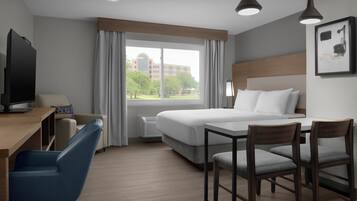 Quarto de hotel com cama, escrivaninha, cadeira e vista para um edifício pela janela.