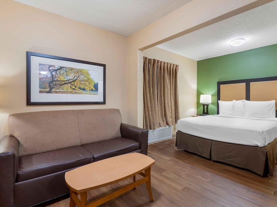 Quarto de hotel com cama, sofá, mesa pequena, quadro na parede e janela com cortinas.