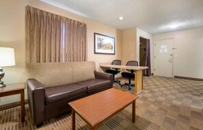 Quarto de hotel com um sofá de couro marrom, uma mesa de centro de madeira, uma escrivaninha com duas cadeiras, um quadro emoldurado na parede e uma porta com interruptor de luz.